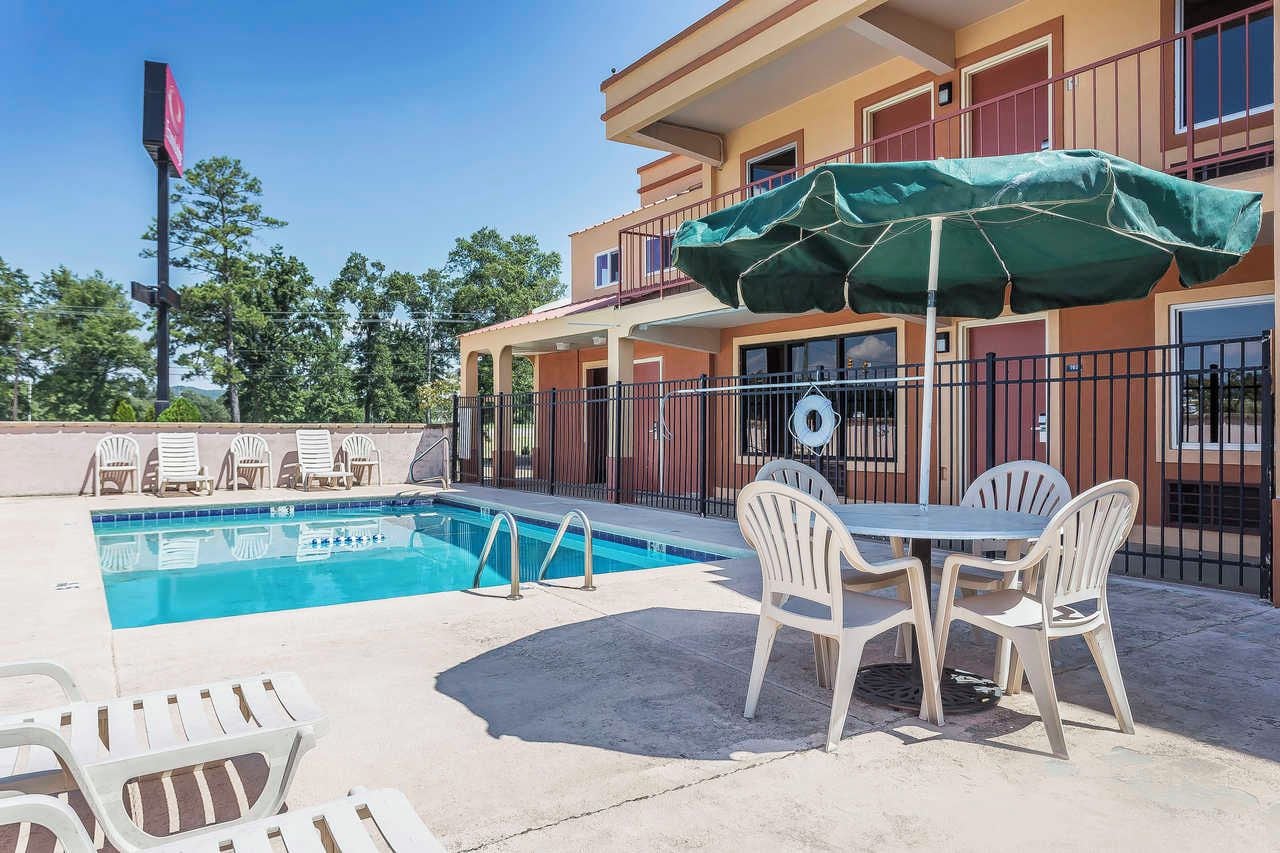 Uma piscina com espreguiçadeiras e uma mesa, rodeada por um edifício com varandas e uma vedação.