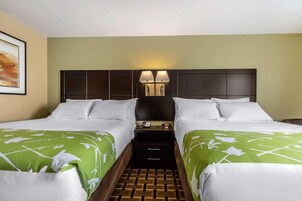 Quarto de hotel com duas camas, cabeceira com luminárias fixadas na parede, mesa de cabeceira com telefone e abajur, e piso estampado.
