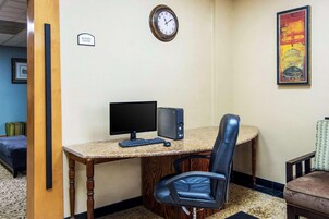 Quarto de hotel com escrivaninha de madeira, computador, cadeira, cama e um quadro na parede.