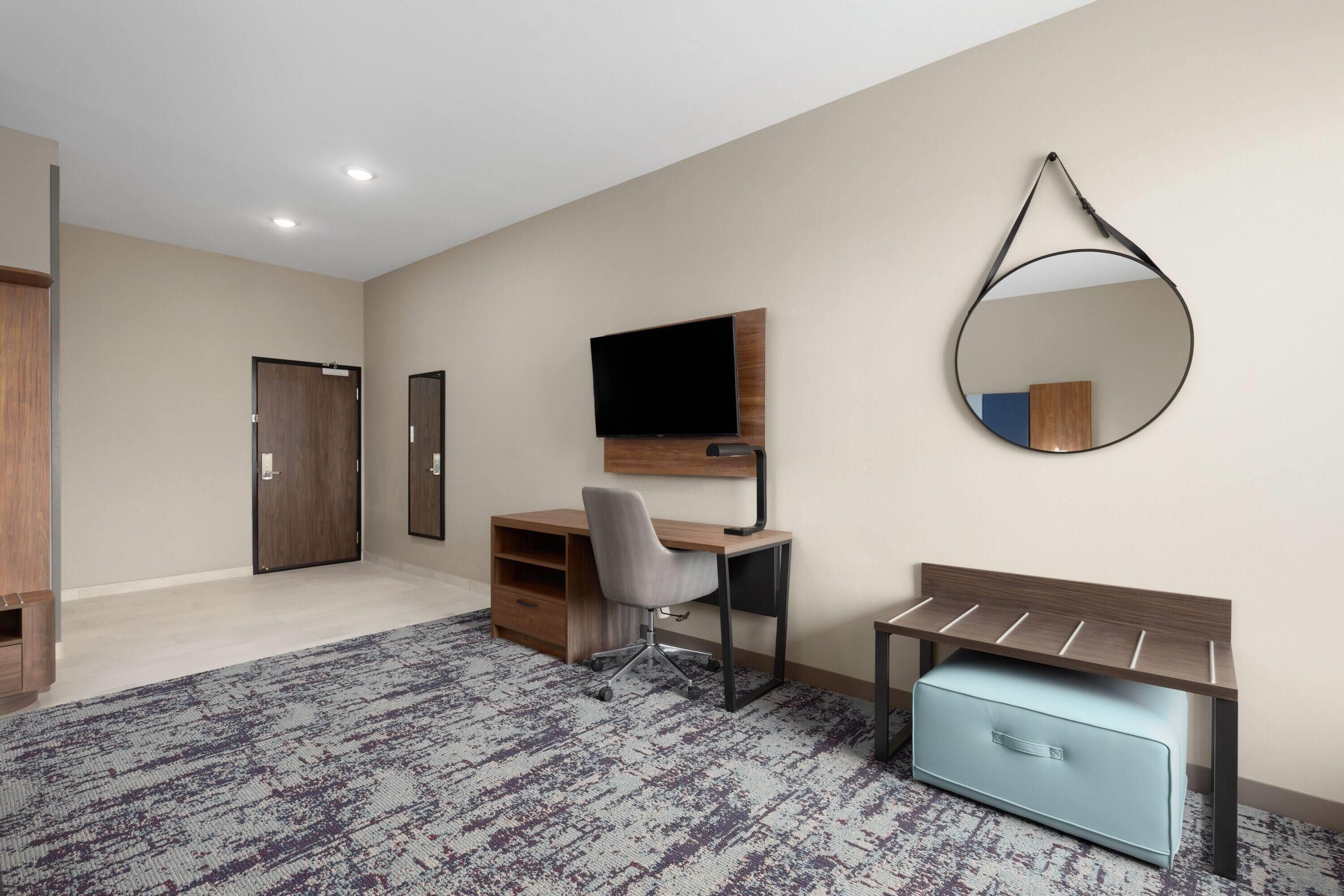 Quarto de hotel com escrivaninha, cadeira, televisão, espelho, guarda-roupa e uma mala azul.