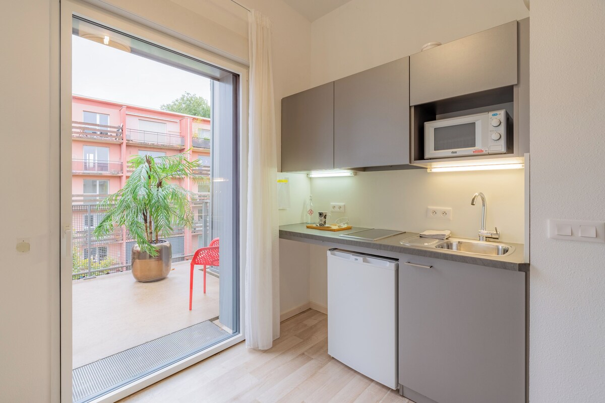 Cozinha compacta com micro-ondas, pia e lava-louças. Uma planta em vaso é visível pela porta de vidro deslizante, e uma cadeira vermelha fica do lado de fora.