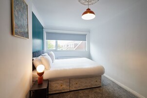 Quarto com cama, mesa de cabeceira, luminária e janela com vista para edifícios.