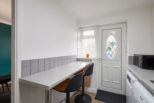 Uma cozinha compacta com bancada branca, dois banquetes pretos, micro-ondas e porta com vitrais.