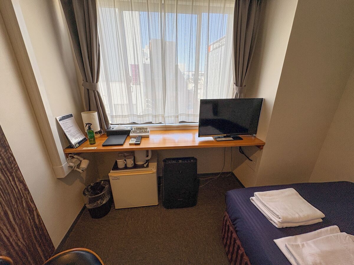 Quarto de hotel com escrivaninha, cama, televisão, mini-geladeira, lixeira e janela com cortinas.