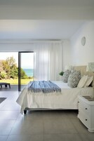 Quarto com uma cama grande, mesa de cabeceira, vista para o oceano e porta de vidro corrediça que dá para uma varanda.