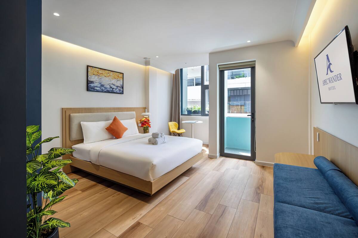 Quarto de hotel moderno com cama, sofá azul, mesa pequena e planta em vaso.