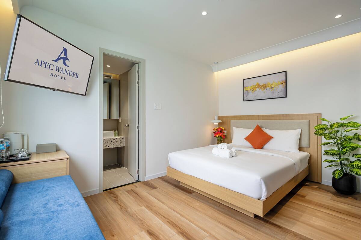 Quarto de hotel com uma cama, mesa de cabeceira, planta em vaso e banheiro visível por uma porta aberta.