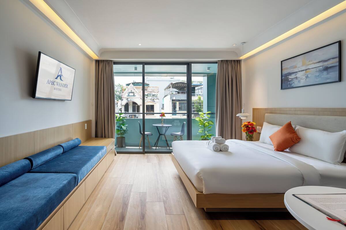 Quarto de hotel com cama, sofá, escrivaninha e vista da cidade através de grandes janelas.