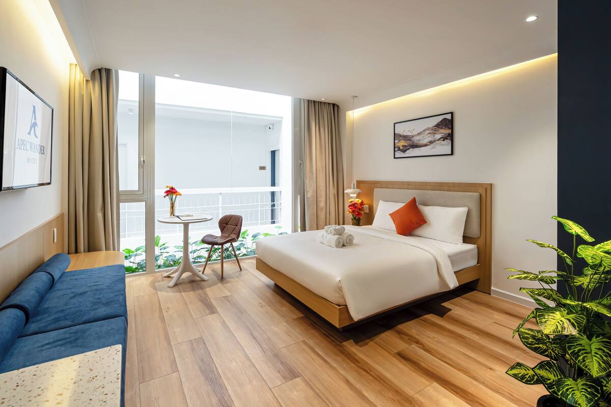 Quarto de hotel moderno com uma cama grande, uma mesinha com uma cadeira, um sofá e uma varanda com plantas.