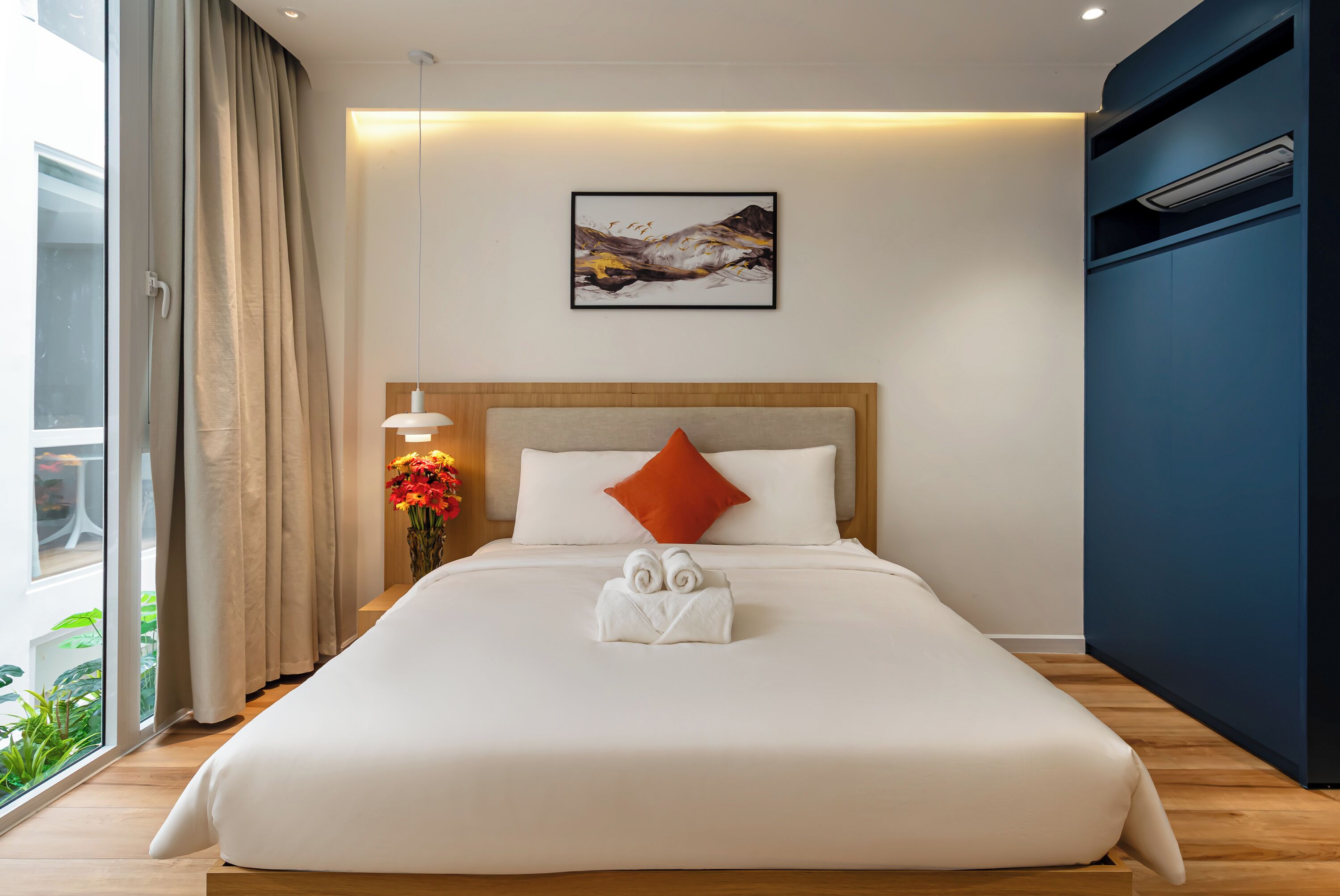 Quarto de hotel com uma cama grande, cabeceira de madeira, travesseiro vermelho, arranjo de toalhas brancas, um quadro na parede e uma janela com cortinas.