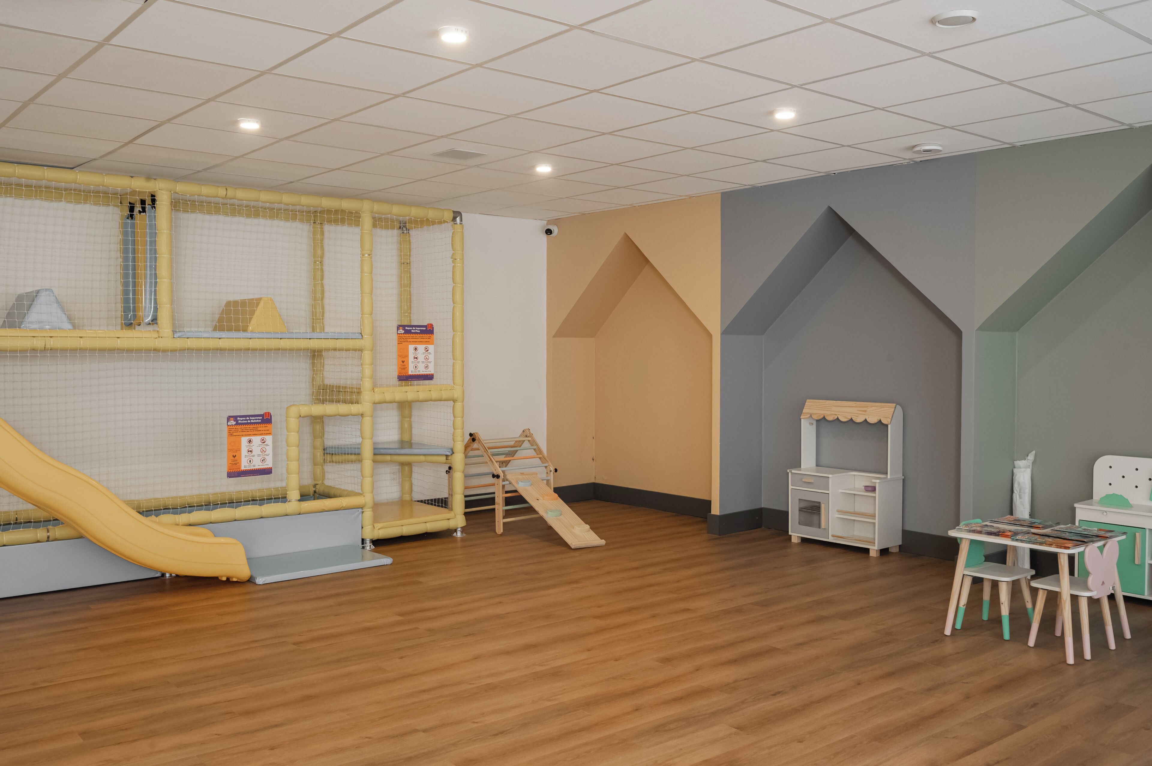 Uma sala de brinquedos com um escorrega, rede de escalada e uma mesa pequena com cadeiras.