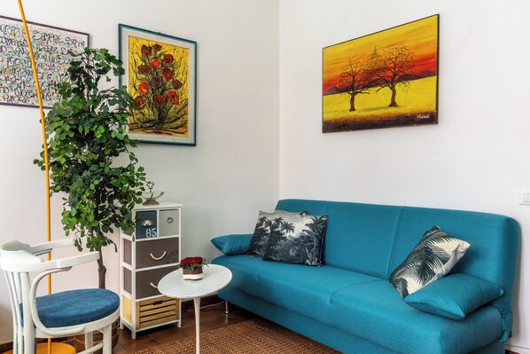 Uma sala de estar com um sofá verde-água, uma cadeira branca, uma mesinha e uma planta. Há duas obras de arte emolduradas na parede.