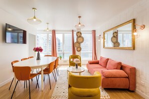 Uma sala de estar moderna com uma mesa de jantar branca, cadeiras laranja, uma poltrona amarela, um sofá rosa e um espelho grande na parede.