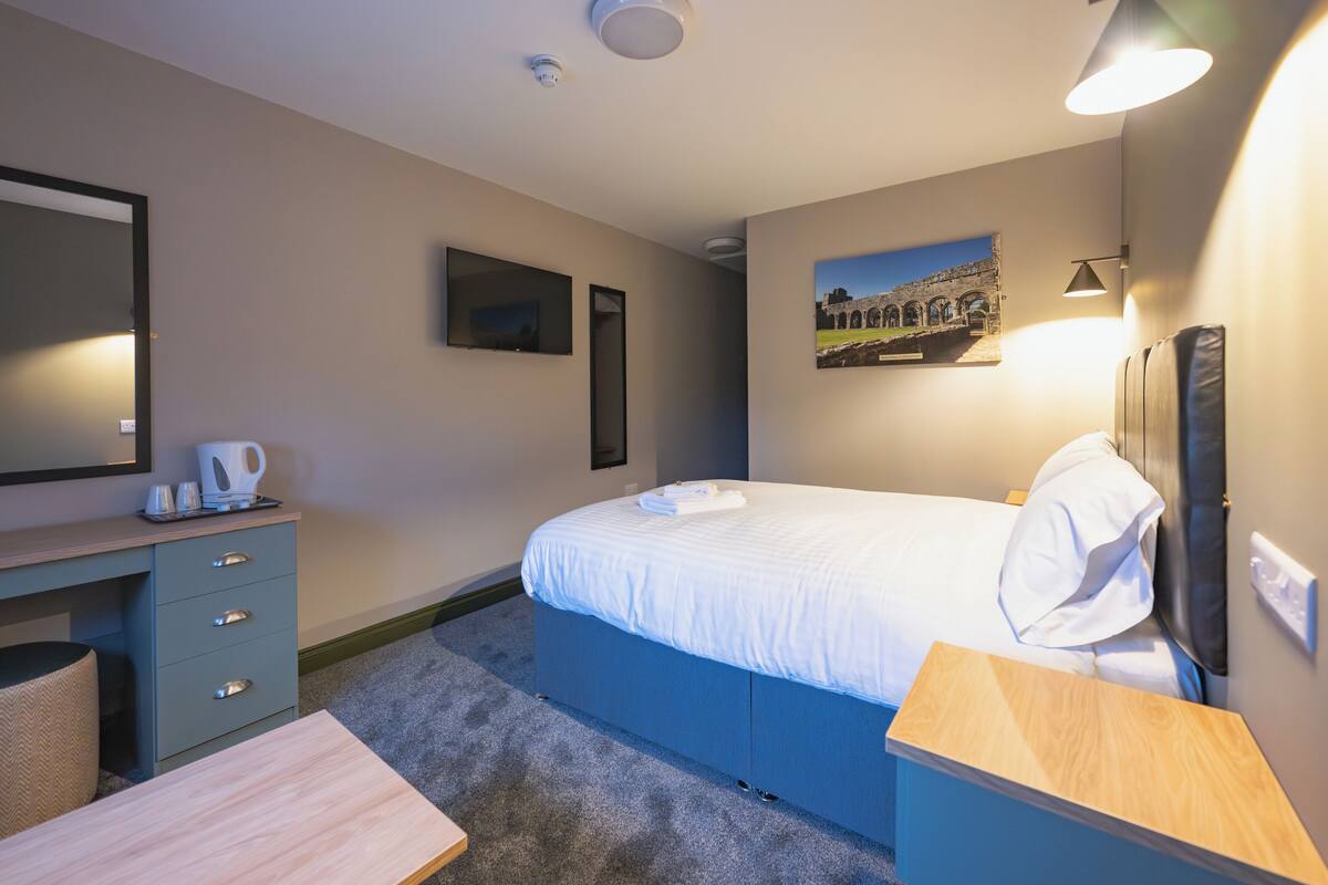 Quarto de hotel com cama, escrivaninha, cadeira, televisão e um quadro na parede.