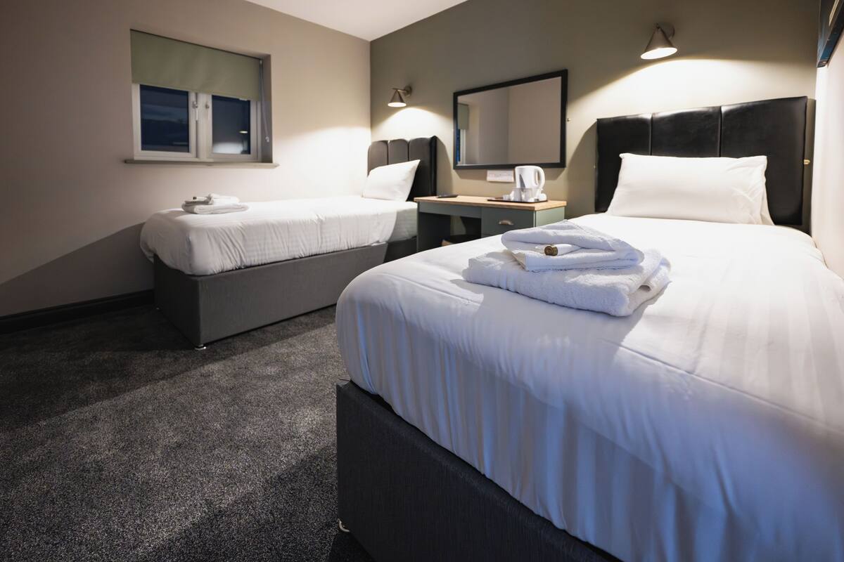 Quarto de hotel com duas camas, uma escrivaninha, uma cafeteira e um espelho.