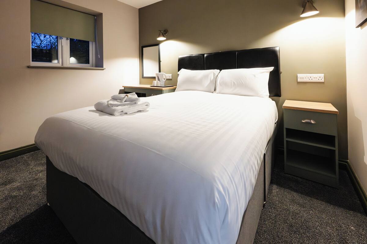 Uma cama bem arrumada com roupa de cama branca, uma mesinha de cabeceira com abajur e uma janela com vista para árvores.