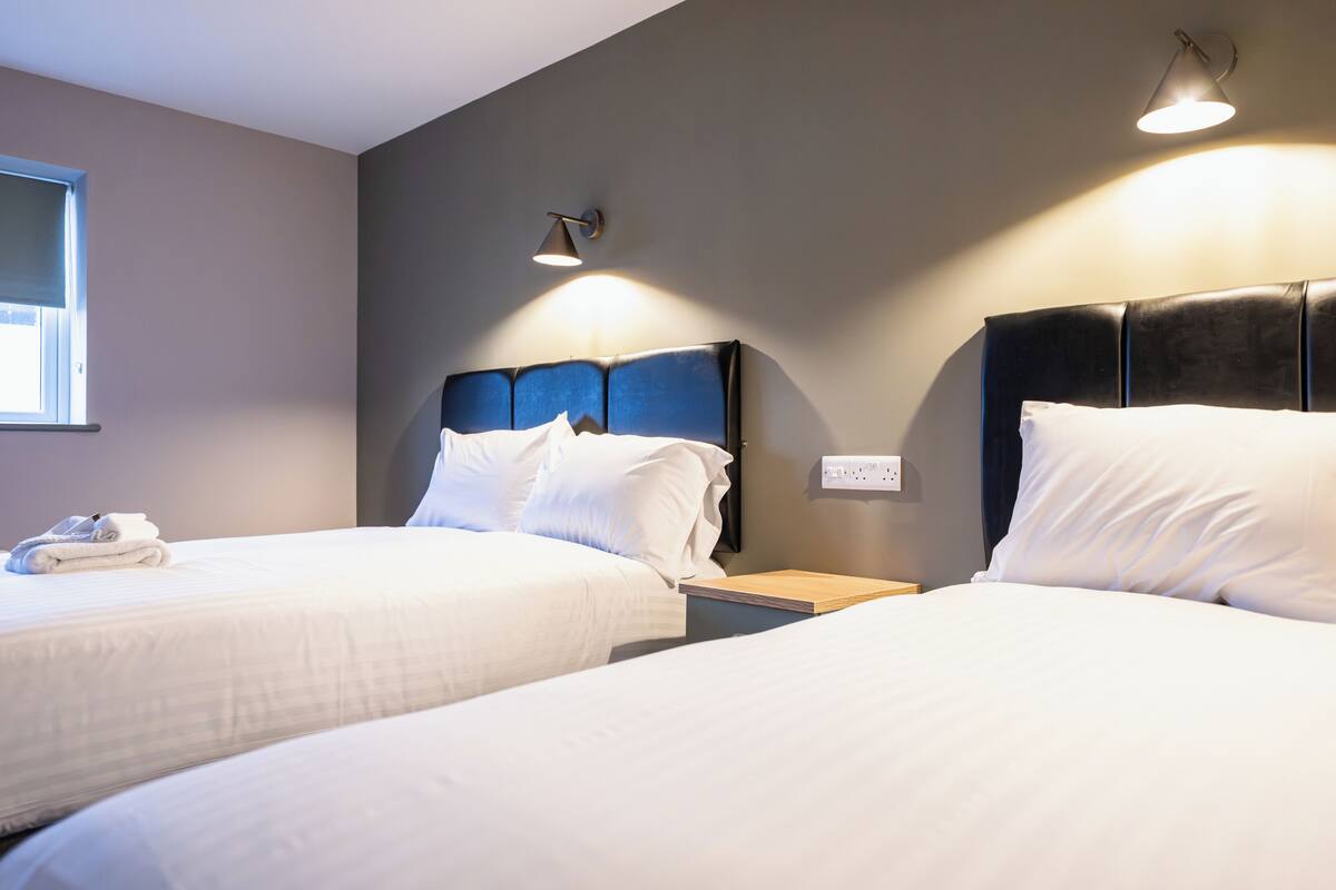 Quarto de hotel com duas camas, cada uma com cabeceira e roupa de cama branca. Uma mesa de cabeceira com luminária e tomada está presente. Uma janela é visível no lado esquerdo do quarto.