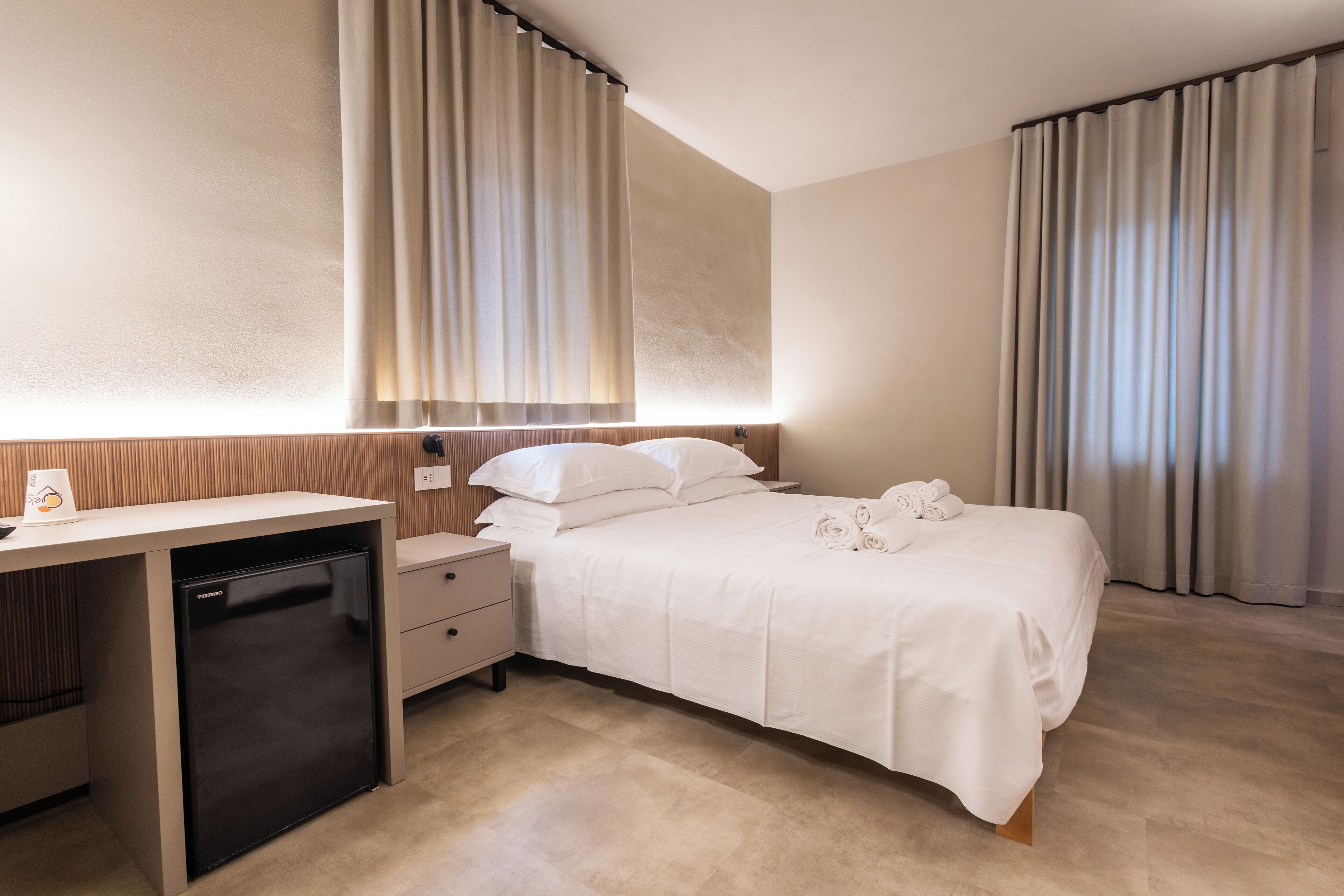 Quarto de hotel com cama, mesa de cabeceira e frigorífico embutido.