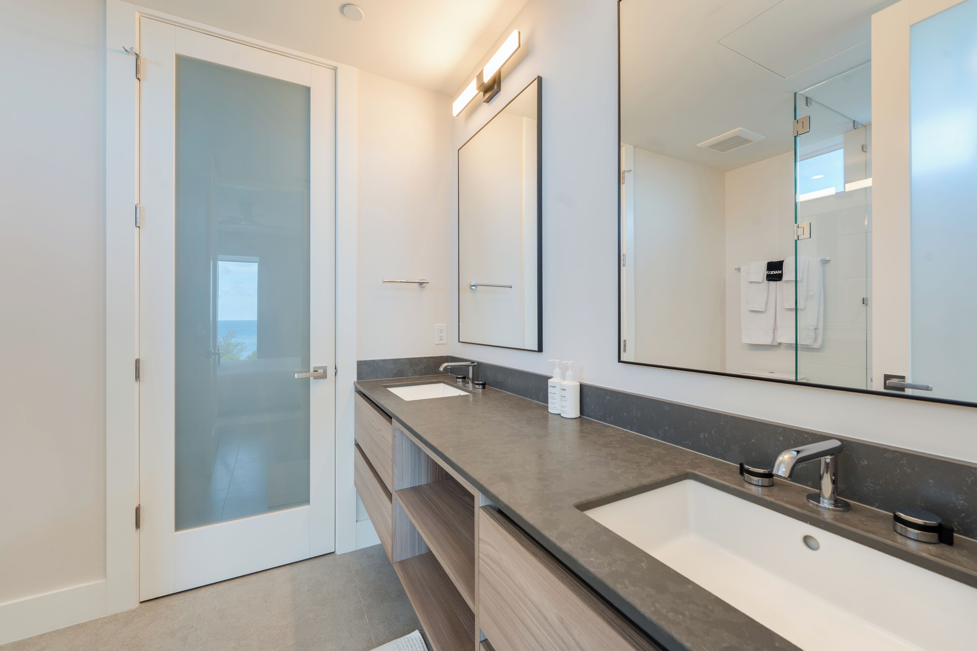 Uma casa de banho moderna com duas pias, um espelho grande e uma porta de vidro fosco.