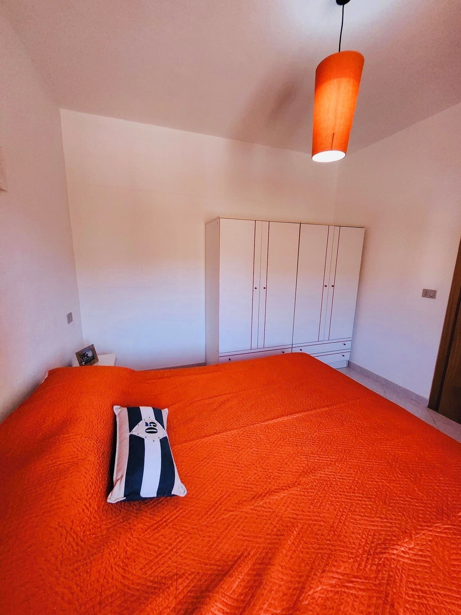 Quarto com cama grande, guarda-roupa branco e luminária suspensa laranja.