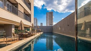 Piscina com espreguiçadeiras e guarda-sóis, cercada por um edifício com varandas.