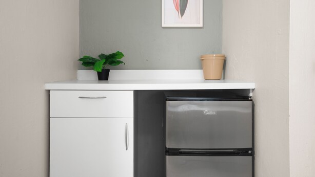Uma cozinha compacta com bancada branca, um pequeno vaso com planta, um cesto de lixo bege e uma geladeira de aço inoxidável.