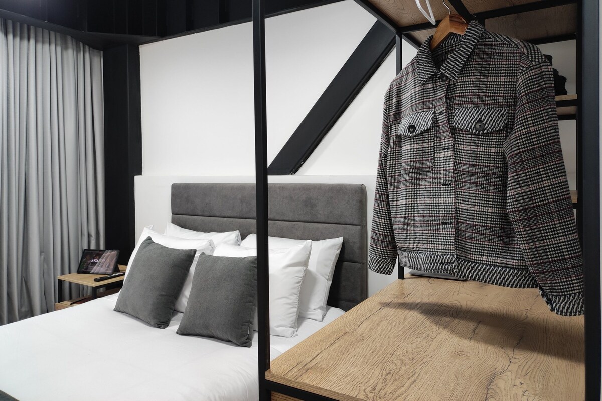 Quarto moderno com uma cama, almofadas cinzentas, uma mesinha de cabeceira de madeira e um cabideiro.