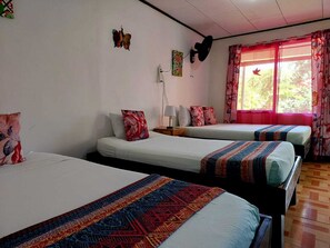 Quarto de hotel com duas camas, uma janela com cortinas, um ventilador fixado na parede e uma pequena mesa com luminária.