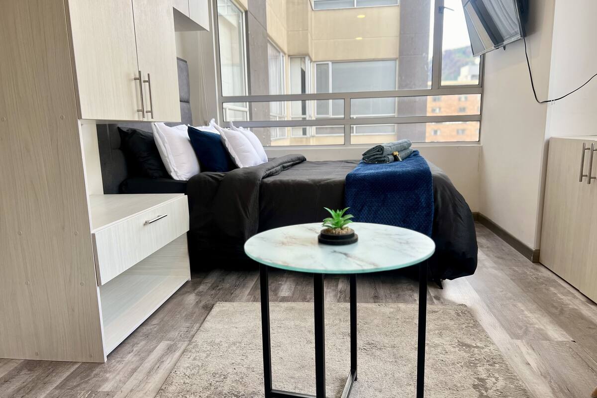 Quarto moderno e compacto, com cama, uma mesinha com uma planta, um sofá e uma janela ampla com vista para a cidade.