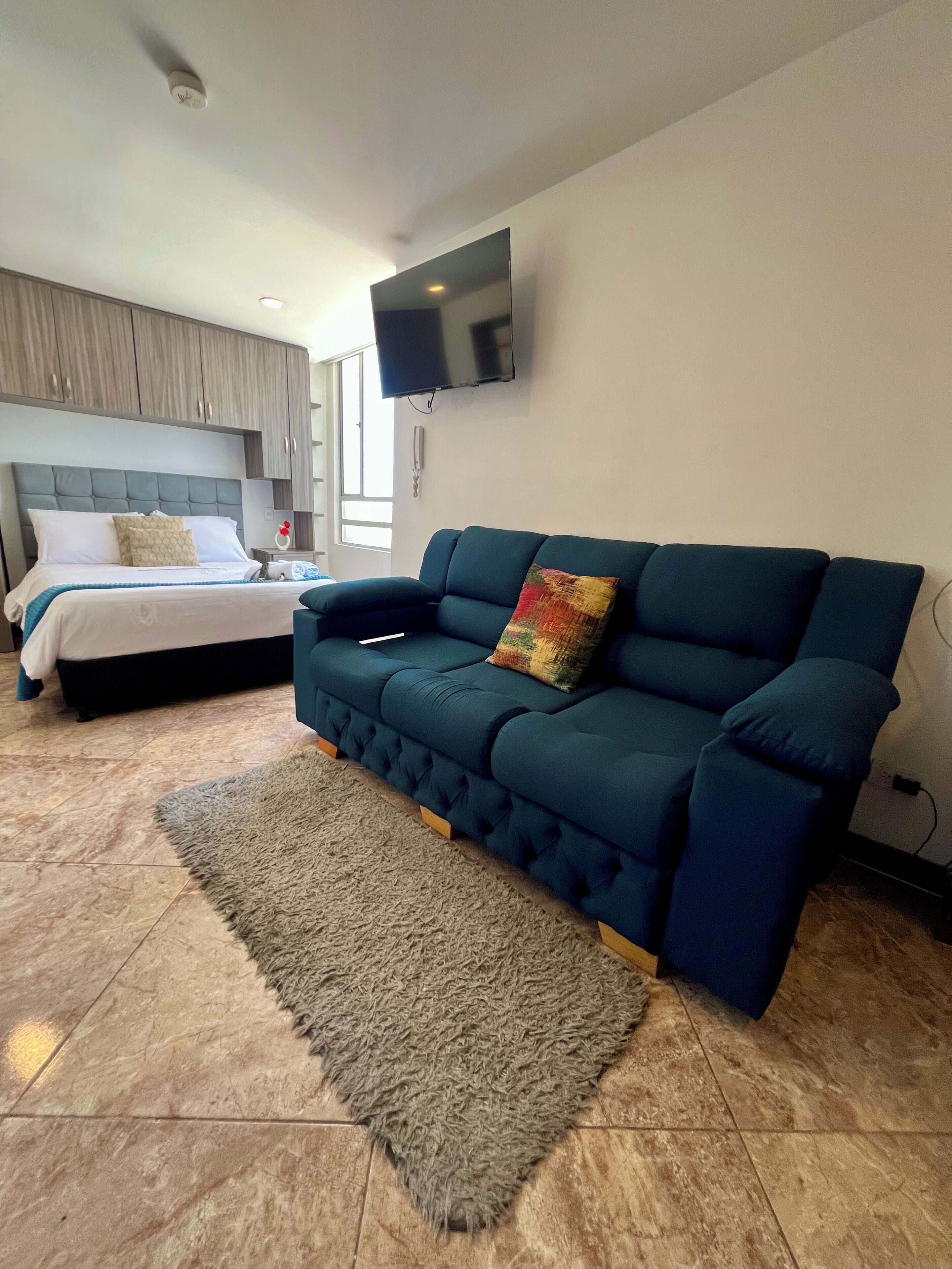 Um quarto moderno com uma cama, um sofá azul, um tapete cinza e uma TV de tela plana fixada na parede.