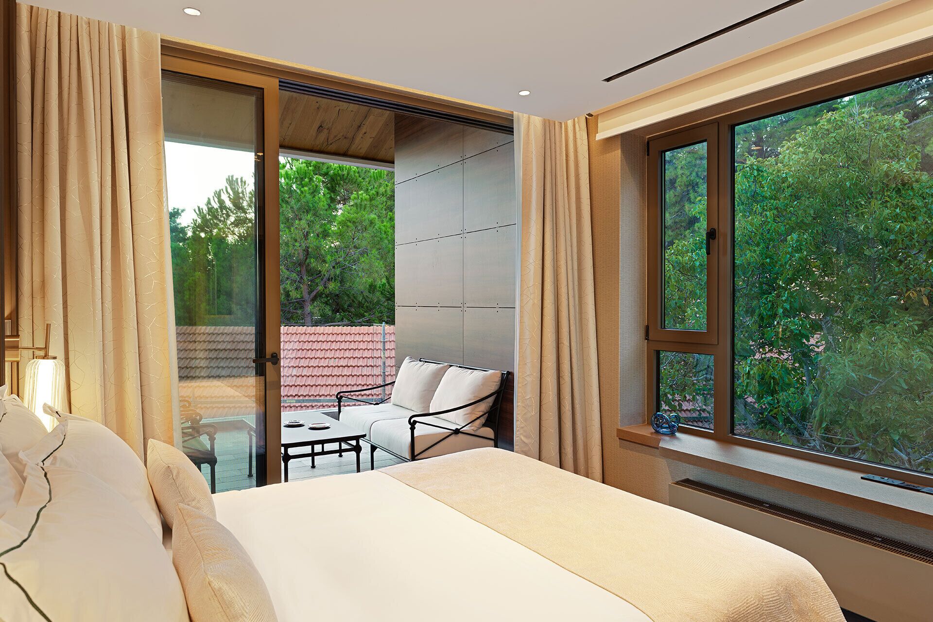Quarto moderno com uma cama grande, uma mesinha e uma cadeira. Tem vista para árvores e um edifício com telhado vermelho lá fora.