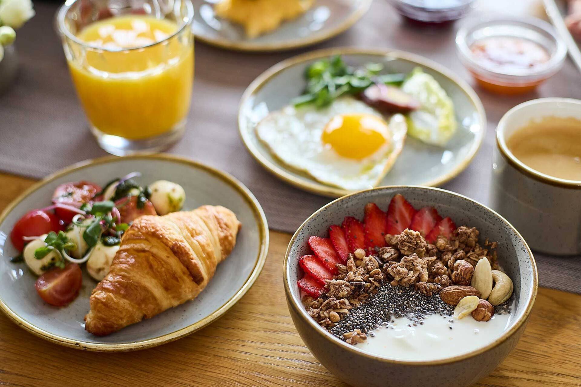Uma mesa de café da manhã com croissants, uma tigela de iogurte com frutas e nozes, um copo de suco de laranja e um prato com ovos e vegetais.