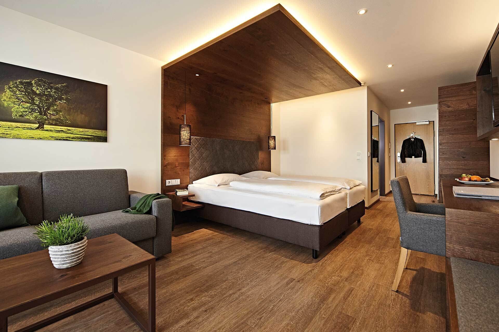 Quarto de hotel moderno com uma cama, um sofá, uma pequena mesa com uma planta e uma pintura de uma árvore.