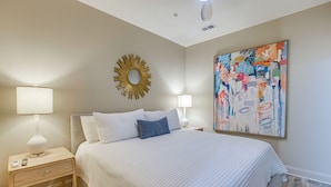 Quarto com uma cama grande, duas mesas de cabeceira com luminárias, um espelho de sol decorativo e uma pintura abstrata colorida na parede.