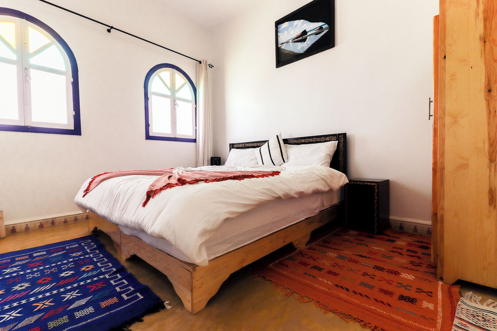 Quarto com cama de madeira, tapete azul e vermelho, parede branca e quadro emoldurado.
