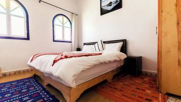 Quarto com cama de madeira, tapete azul e vermelho, parede branca e quadro emoldurado.