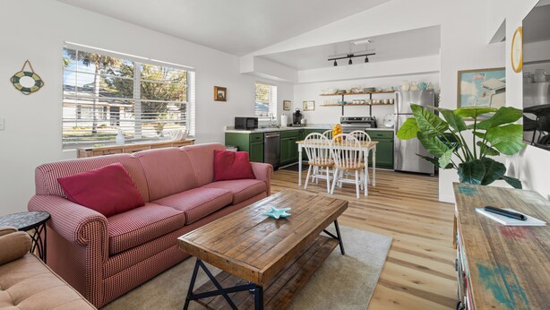 Uma sala de estar ampla com um sofá xadrez vermelho e branco, uma mesa de centro de madeira e uma mesa de jantar com cadeiras. Uma planta grande está no canto, e a cozinha é visível ao fundo.