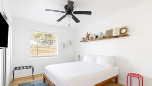 Quarto com cama, ventilador de teto, janela com persianas, piso de madeira, tapete azul, um banquinho vermelho e uma estante com objetos decorativos.