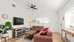 Uma sala de estar com piso de madeira, um sofá marrom, um sofá xadrez vermelho, uma mesa de centro, uma TV, um ventilador de teto, uma janela com persianas, um vaso de planta e um armário de madeira.