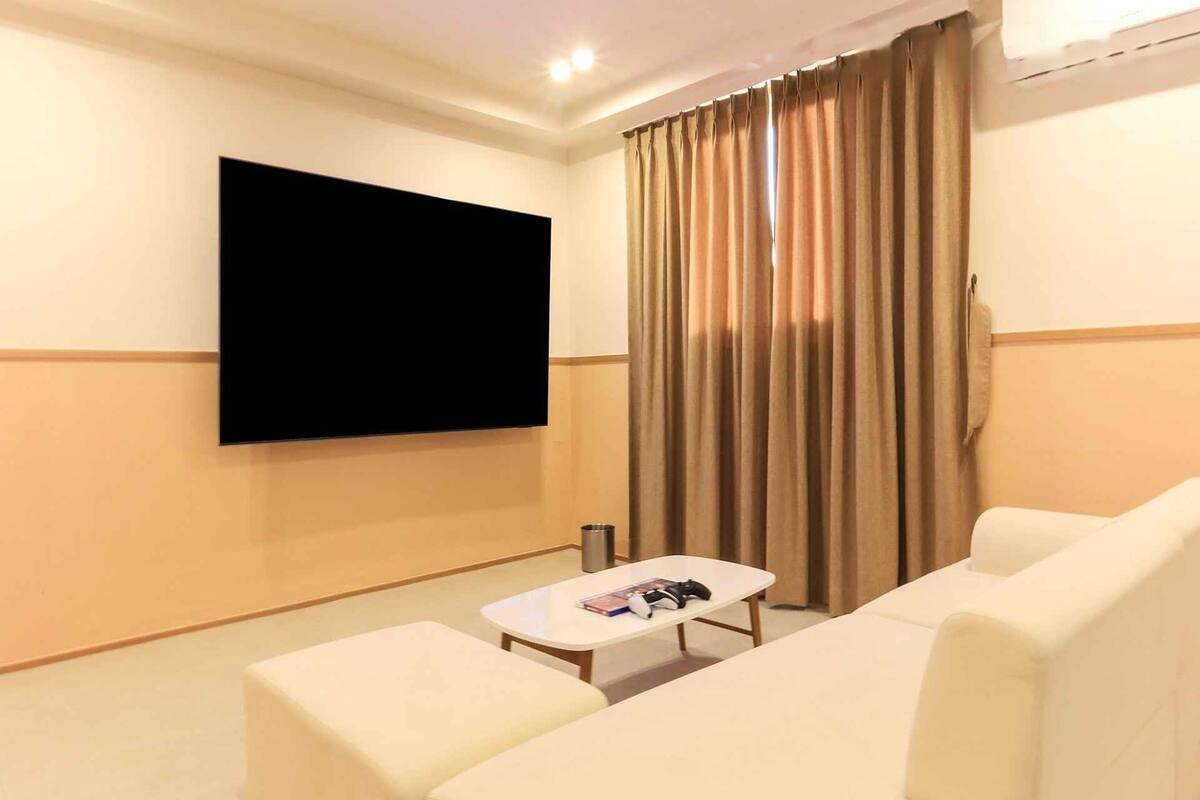 Quarto de hotel com televisão de ecrã plano na parede, um sofá branco, uma pequena mesa branca com um comando remoto e uma janela com cortinas bege.