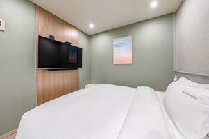 Quarto de hotel com cama, televisão fixada em um painel de madeira e um quadro na parede.