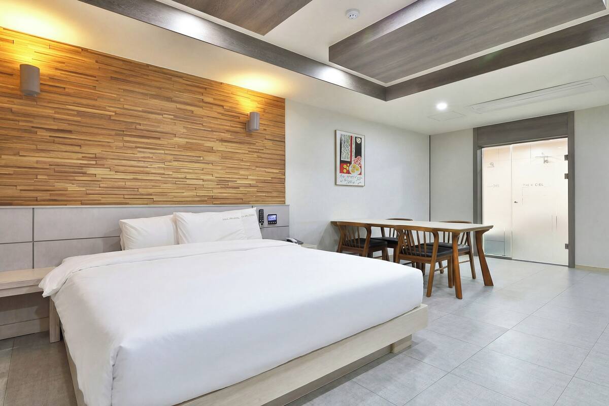 Quarto de hotel moderno com uma cama grande, painel de parede de madeira, uma mesa de jantar e um banheiro visível por uma porta aberta.