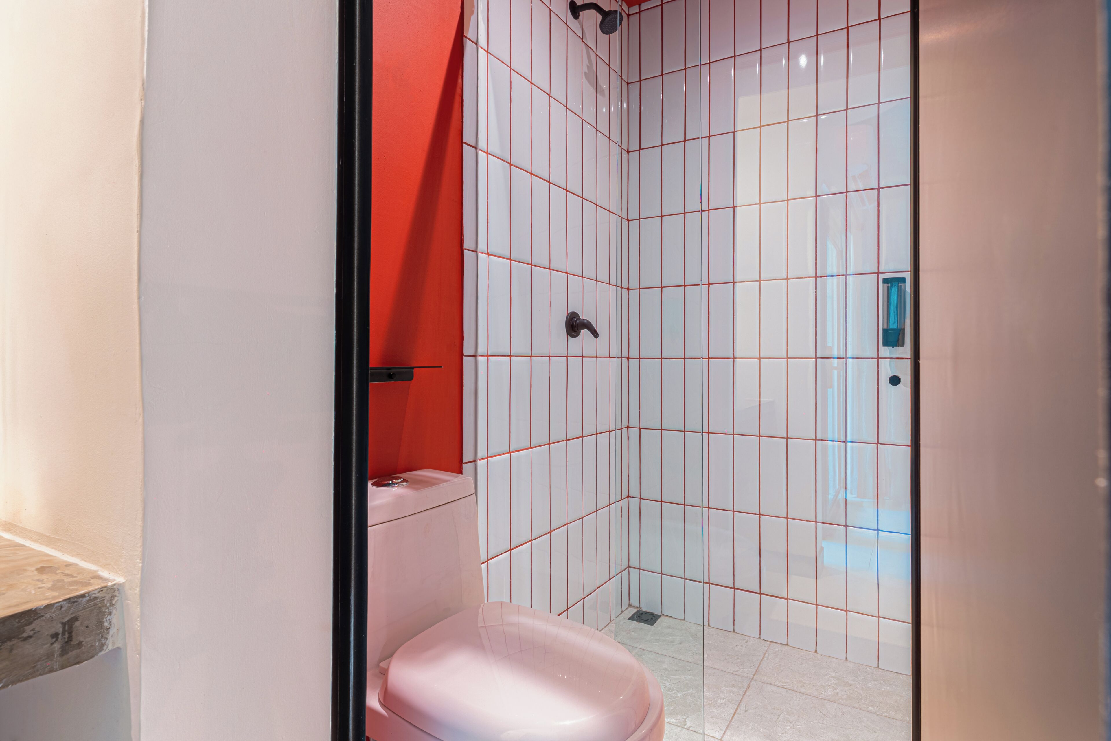 Um banheiro com chuveiro revestido de azulejos brancos, vaso sanitário rosa e parede com detalhes em vermelho.