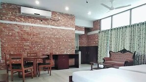 Quarto de hotel com parede de tijolos, uma cama, um sofá, uma escrivaninha com cadeiras e uma janela com cortinas.