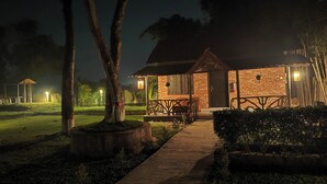 Área externa bem iluminada com um caminho que leva a um edifício de fachada em tijolo e telhado de palha.