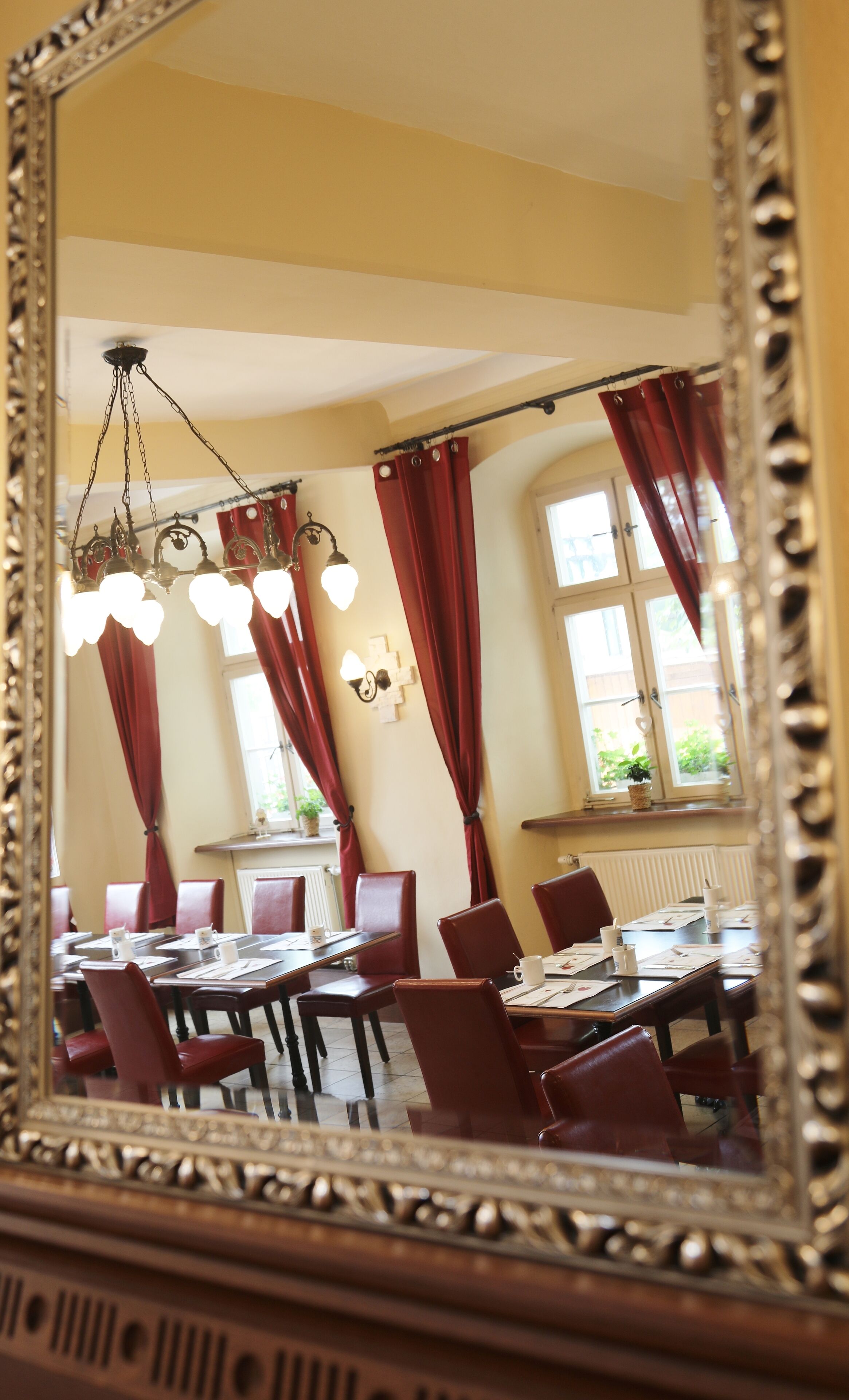 Uma sala de jantar com mesas, cadeiras, um candelabro e cortinas vermelhas.