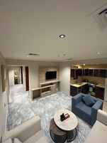 Uma sala de estar moderna com televisão de tela plana, mesa de centro e sofá. Há também uma área de cozinha com armários e bancada.
