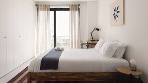 Quarto moderno com cama de madeira, roupa de cama branca, uma escrivaninha com abajur e vista pela janela grande com cortinas.