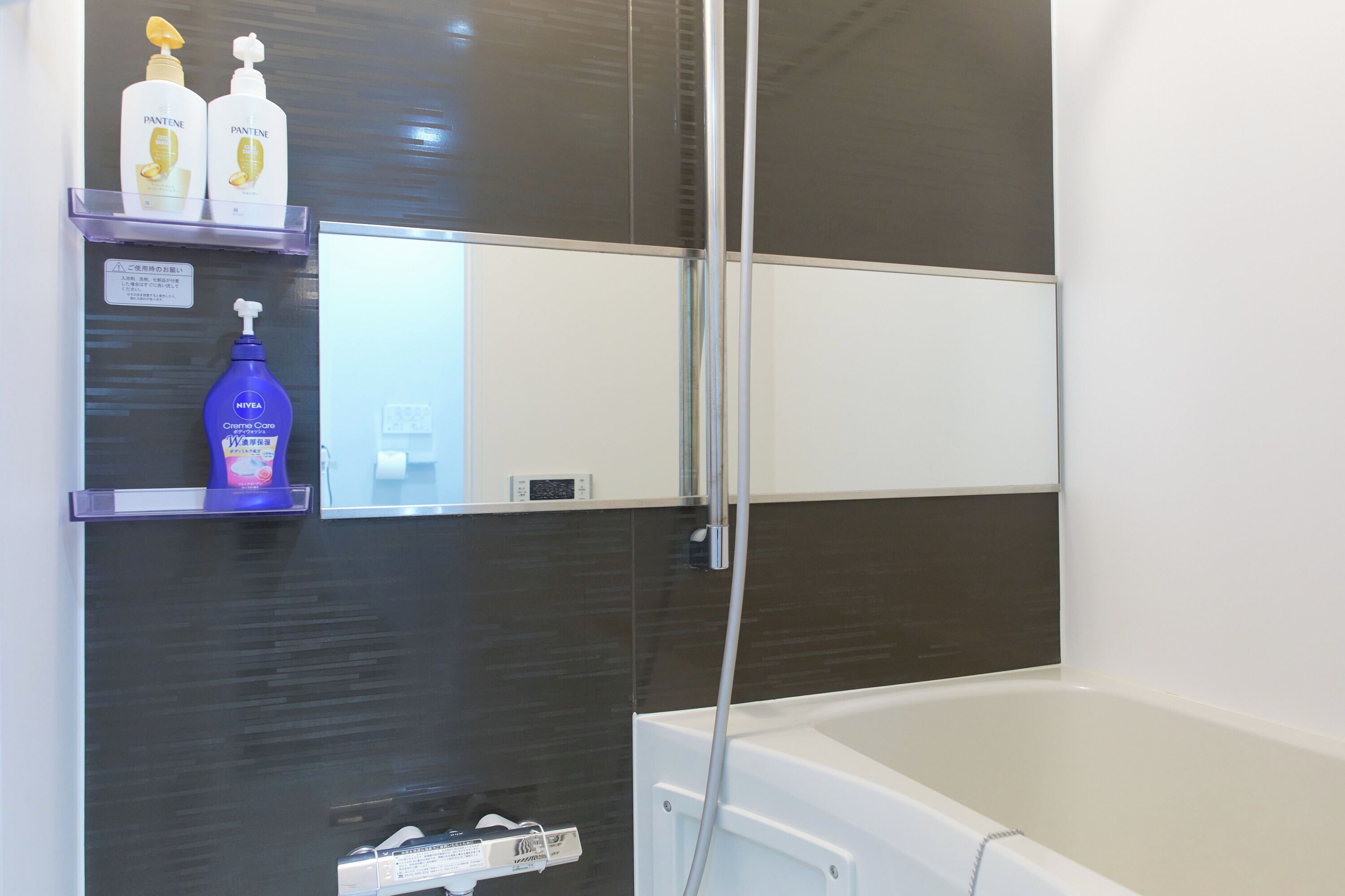 Casa de banho com banheira, um prateleira com produtos Pantene e Nivea, e um espelho.