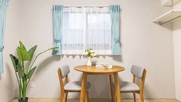 Quarto com cama, mesa redonda de madeira com duas cadeiras, planta em vaso e janela com cortinas.